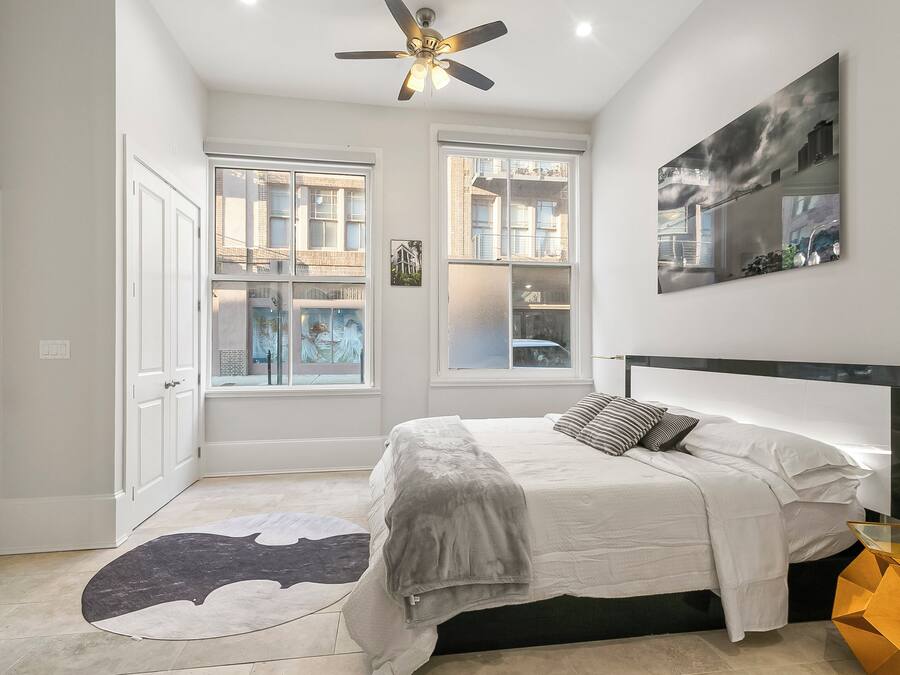 Quarto com cama, ventoinha no teto, duas janelas e tapete.