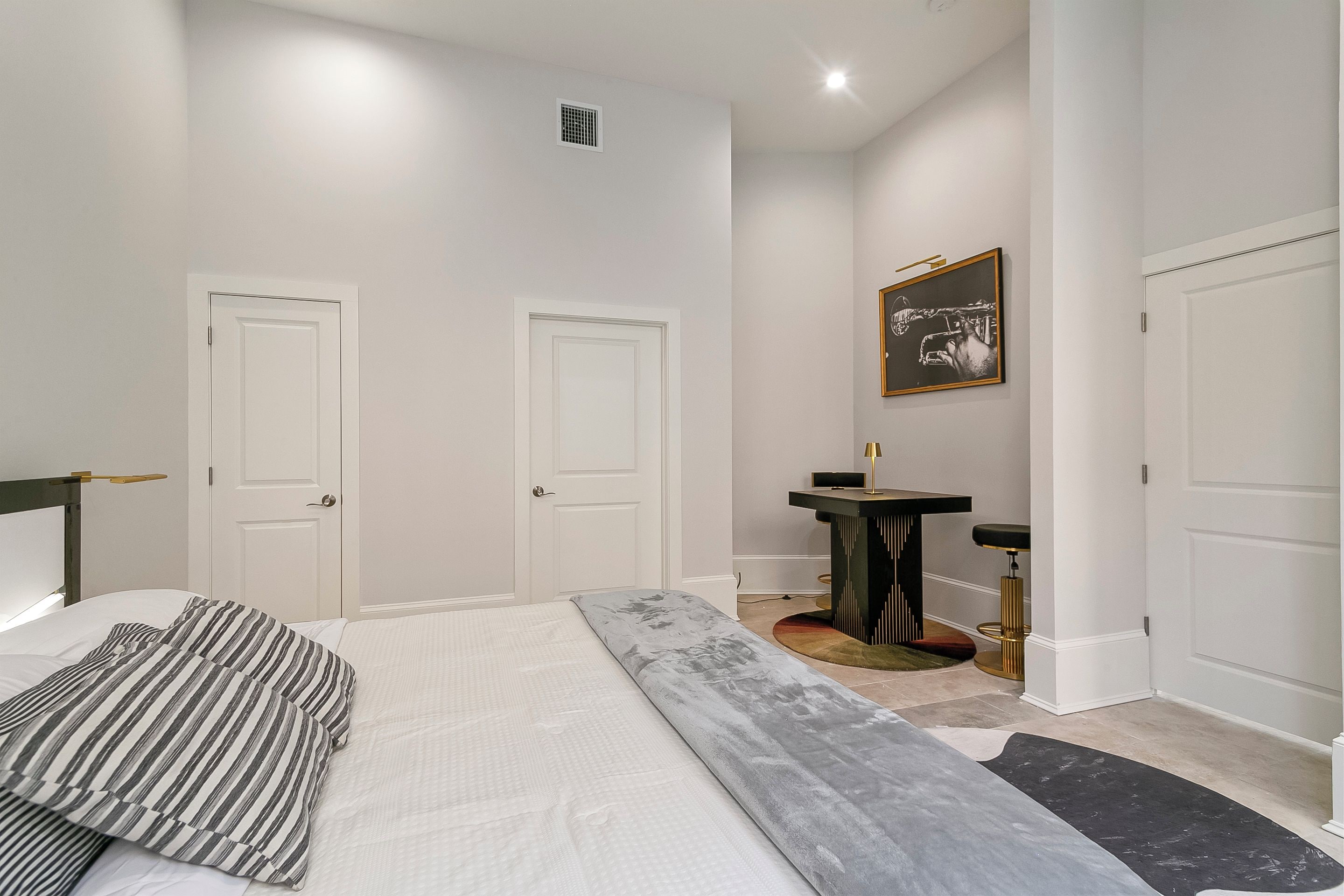 Quarto com cama, duas portas, mesa de cabeceira, uma cadeira, uma mesinha com abajur e um quadro pendurado na parede.