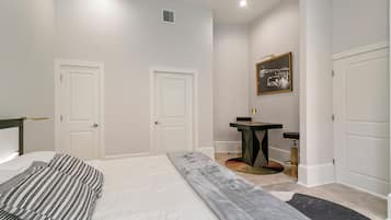 Quarto com cama, duas portas, mesa de cabeceira, uma cadeira, uma mesinha com abajur e um quadro pendurado na parede.