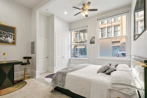 Quarto com cama, escrivaninha, cadeira, ventilador de teto e janela com vista para prédios.