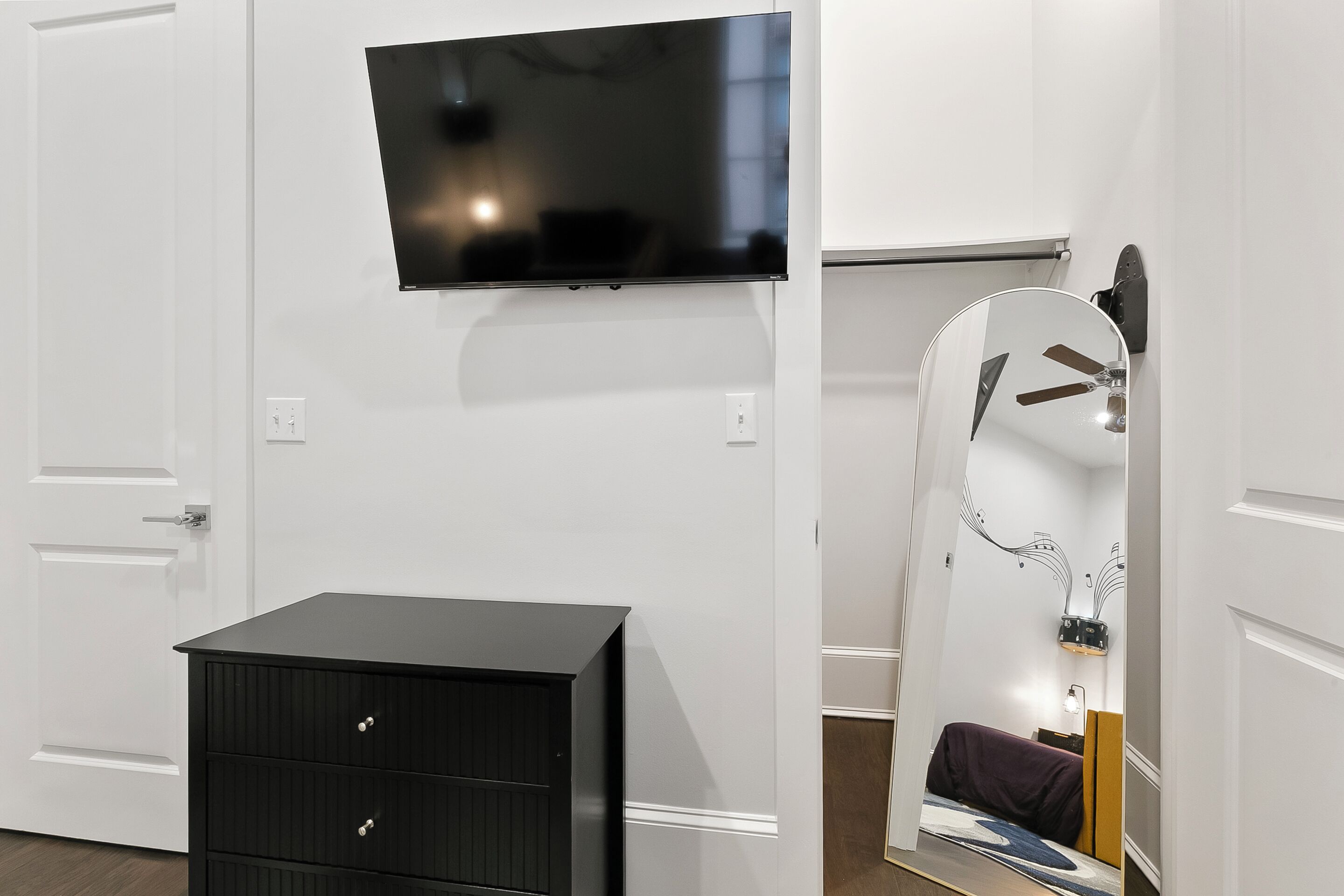 Quarto com televisão de tela plana na parede, cômoda preta com gavetas e um espelho grande com moldura decorativa.