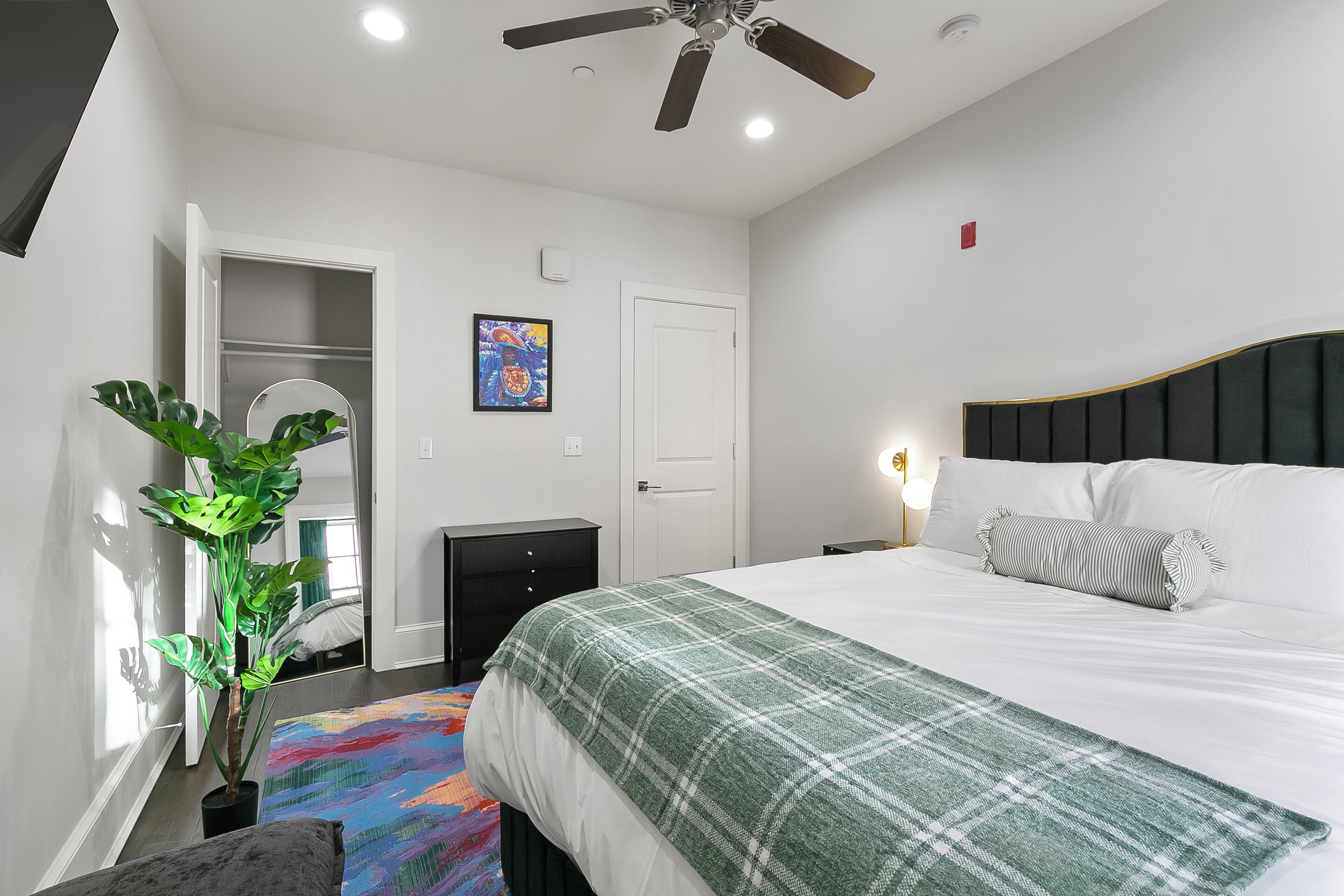 Quarto com cama, ventilador de teto, mesa de cabeceira, abajur, planta e tapete colorido.