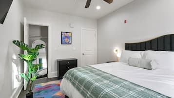 Quarto com cama, ventilador de teto, mesa de cabeceira, abajur, planta e tapete colorido.