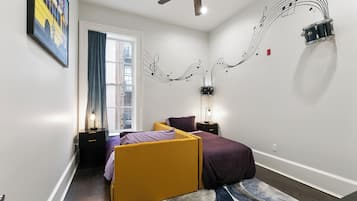 Um quarto com cama, mesa de cabeceira, relógio na parede, ventilador de teto e janela com cortinas.