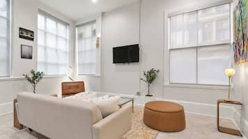 Quarto moderno com cama, mesa de cabeceira, televisão e duas janelas.