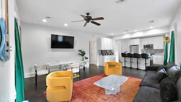 Uma sala de estar moderna com um sofá, cadeiras, uma mesa de centro, televisão e uma área de cozinha com armários e eletrodomésticos.