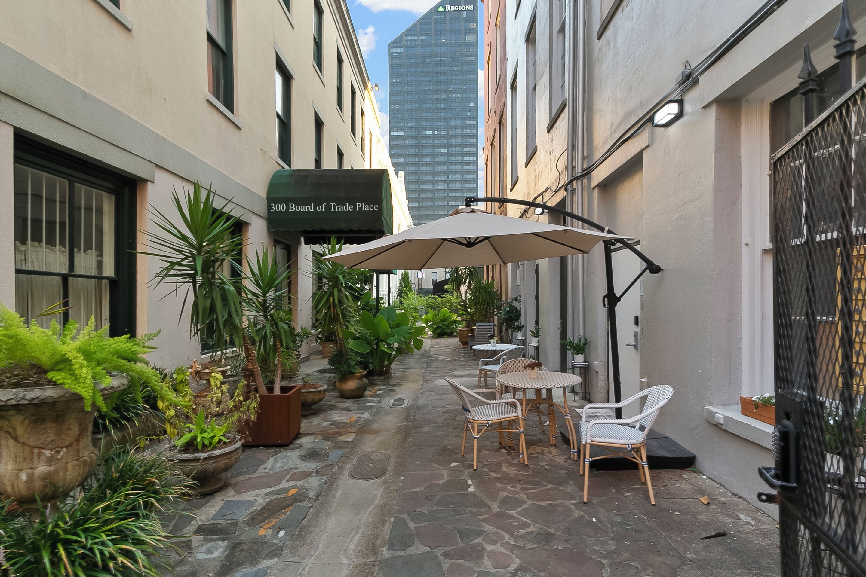Uma viela estreita com mesas ao ar livre, plantas em vasos e um edifício com uma placa indicando '300 Board of Trade Place'.