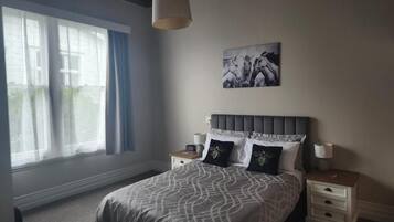 Quarto com cama, mesas de cabeceira, janela com cortinas e uma obra de arte na parede.