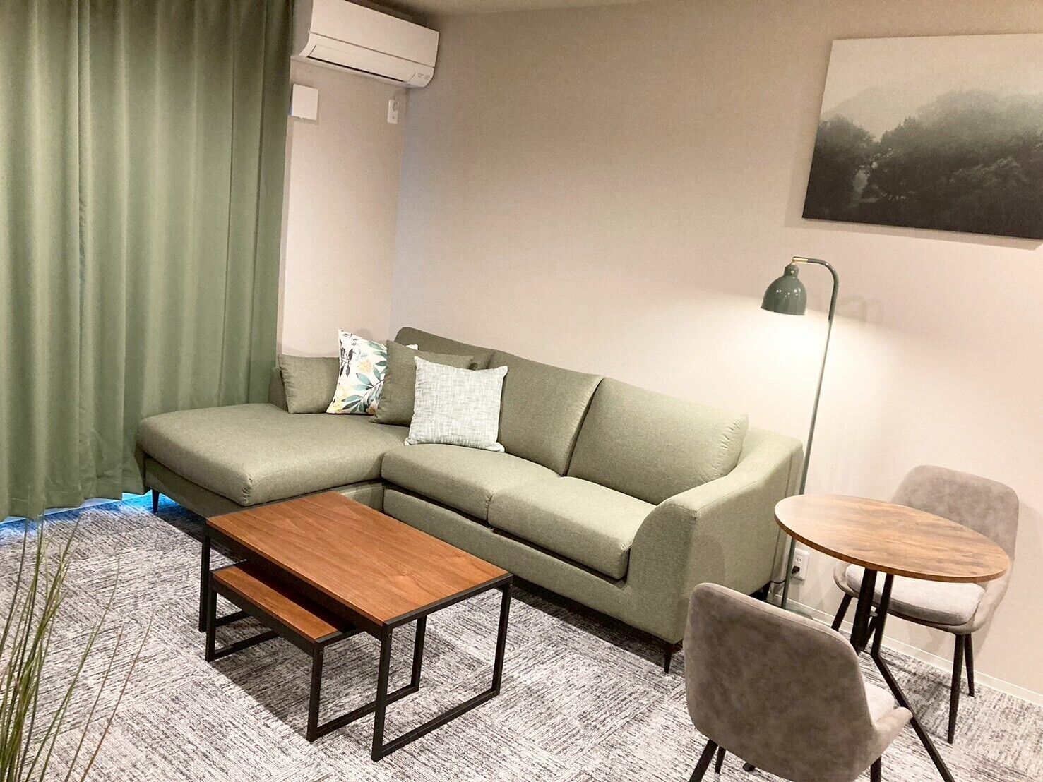 Uma sala de estar com sofá seccional, mesa de centro de madeira, mesa lateral redonda e uma poltrona cinza.