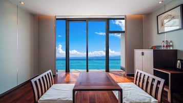 Quarto com uma grande porta de vidro deslizante que dá para uma varanda com vista para o oceano. Há uma mesa de madeira com duas cadeiras e um armário com garrafas em cima.