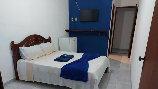 Uma cama de solteiro com colcha branca e azul, uma pequena mesa de cabeceira, uma televisão fixada na parede e uma porta que dá para outro quarto.