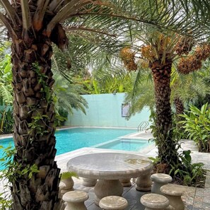 Uma área de piscina com mesa de pedra e bancos, rodeada por palmeiras e plantas tropicais.