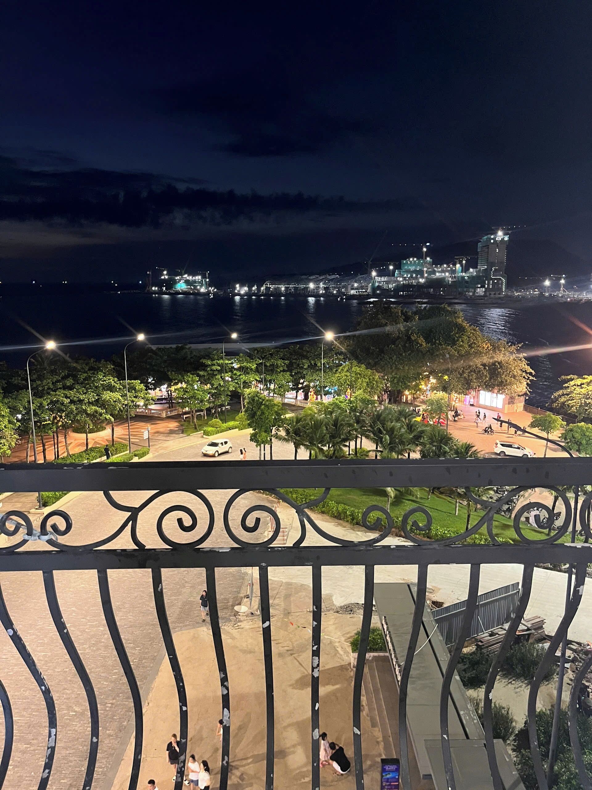 Vista noturna de uma varanda com vista para uma orla iluminada, com palmeiras e um horizonte urbano distante.