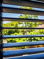 Uma janela com venezianas horizontais, vista para árvores verdes e um prédio ao fundo.