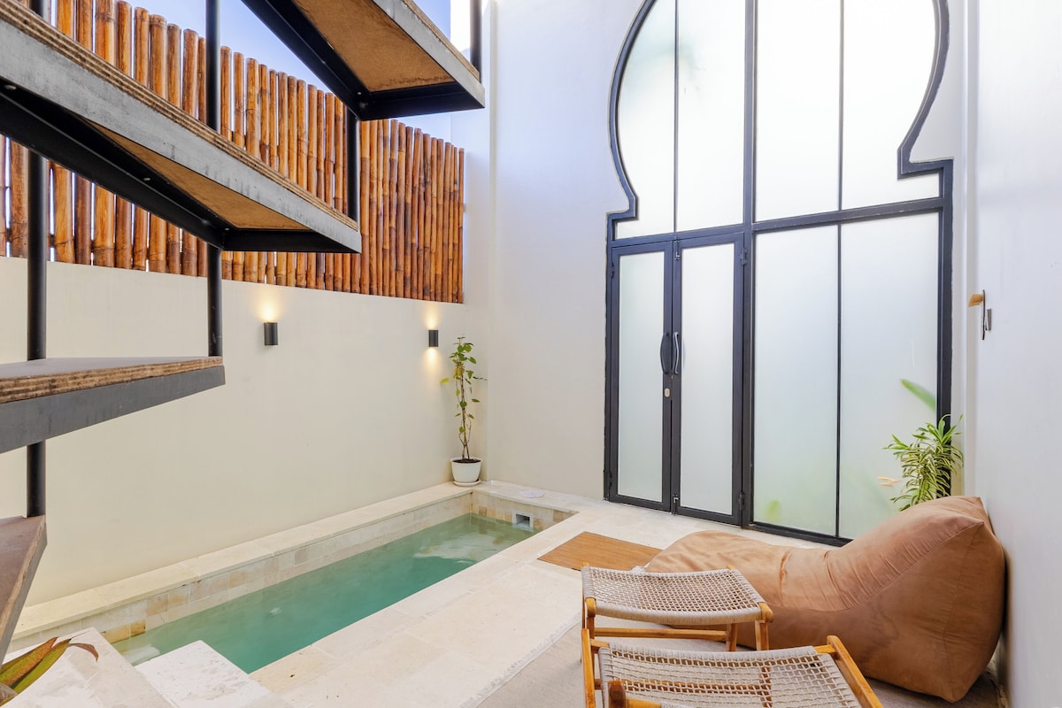 Uma área de piscina com parede de bambu, porta de vidro deslizante e espaço de estar com almofada e cadeira.
