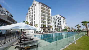 Uma área à beira da piscina com espreguiçadeiras e guarda-sóis, cercada por edifícios altos.