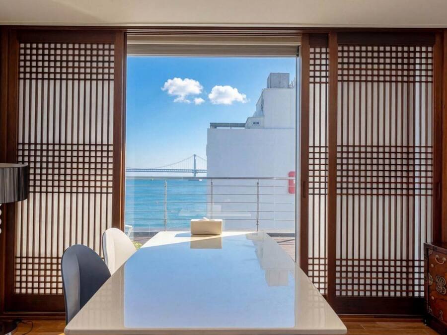 Um quarto com uma mesa grande, cadeiras, um abajur e vista para uma ponte e para a água.