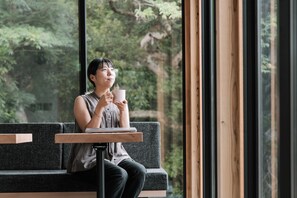 Uma pessoa sentada num banco com um laptop e uma chávena, num espaço com grandes janelas e vista para árvores.