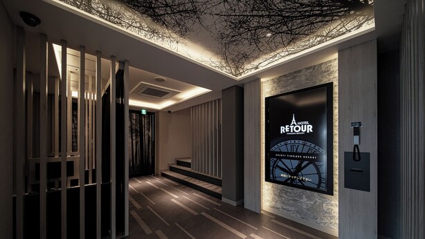Um corredor moderno de hotel com uma tela digital exibindo os textos 'RETOUR' e 'TIMELESS RESORT', uma escadaria e um piso estampado.