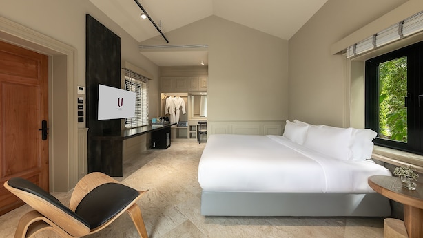 Quarto de hotel com uma cama grande, uma cadeira de madeira, uma escrivaninha com um computador e uma janela com vista para o verde.