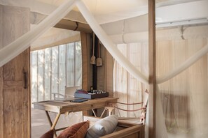 Uma cama de madeira com dossel, uma mesinha com uma caixa e um tablet, e uma janela com cortinas.