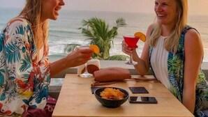 Duas pessoas sentadas numa mesa de madeira com vista para o oceano, uma tigela de petiscos e um smartphone sobre a mesa.
