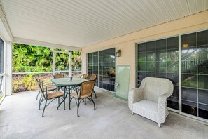 Um alpendre coberto com mesa e cadeiras, um sofá de vime e amplas janelas com vista para o verde.