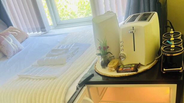 Quarto com cama, mesa de cabeceira com torradeira, pequena mesa com planta e geladeira aberta.