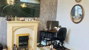 Uma sala de estar com lareira, televisão fixada na parede, escrivaninha com computador e mesa de jogos.