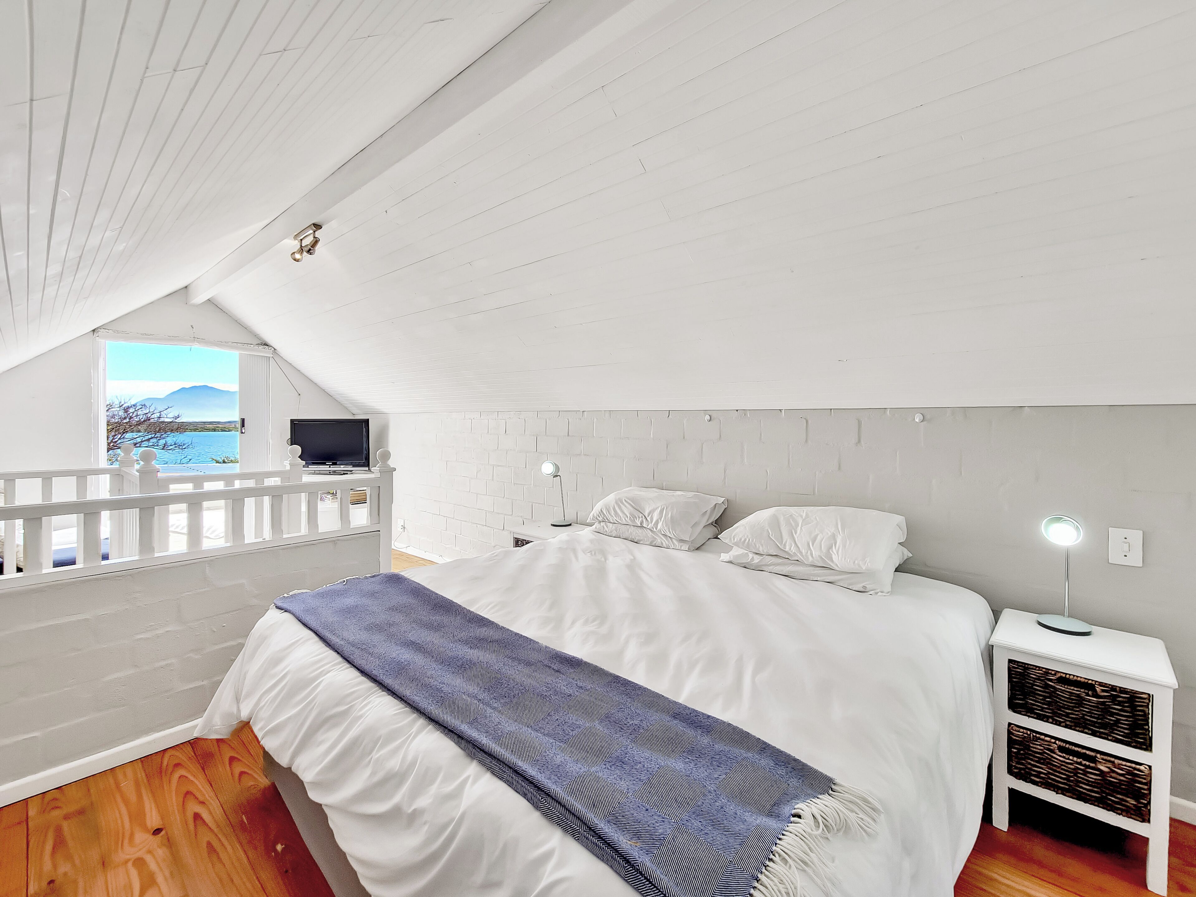Quarto com cama, paredes brancas, chão de madeira, varanda e janela com vista.
