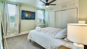 Um quarto com cama, ventilador de teto, televisão e janela com cortinas.