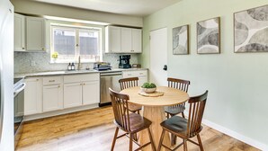 Uma cozinha com armários brancos, uma mesa de jantar de madeira com cadeiras, uma cafeteira e uma janela com vista para uma cerca.