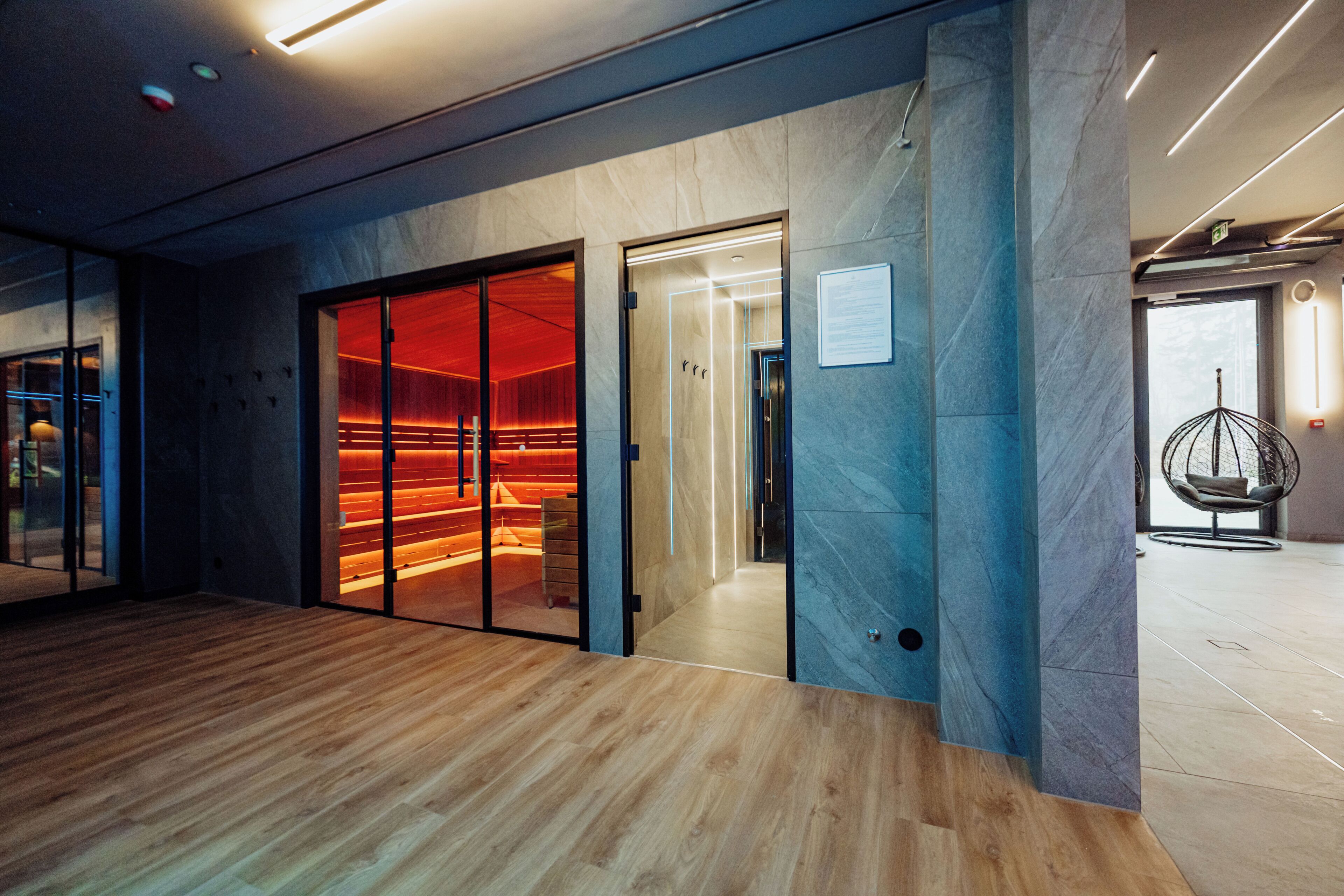 Um interior moderno com piso de madeira, uma porta de vidro que dá para um ambiente iluminado em vermelho e uma cadeira suspensa ao fundo.