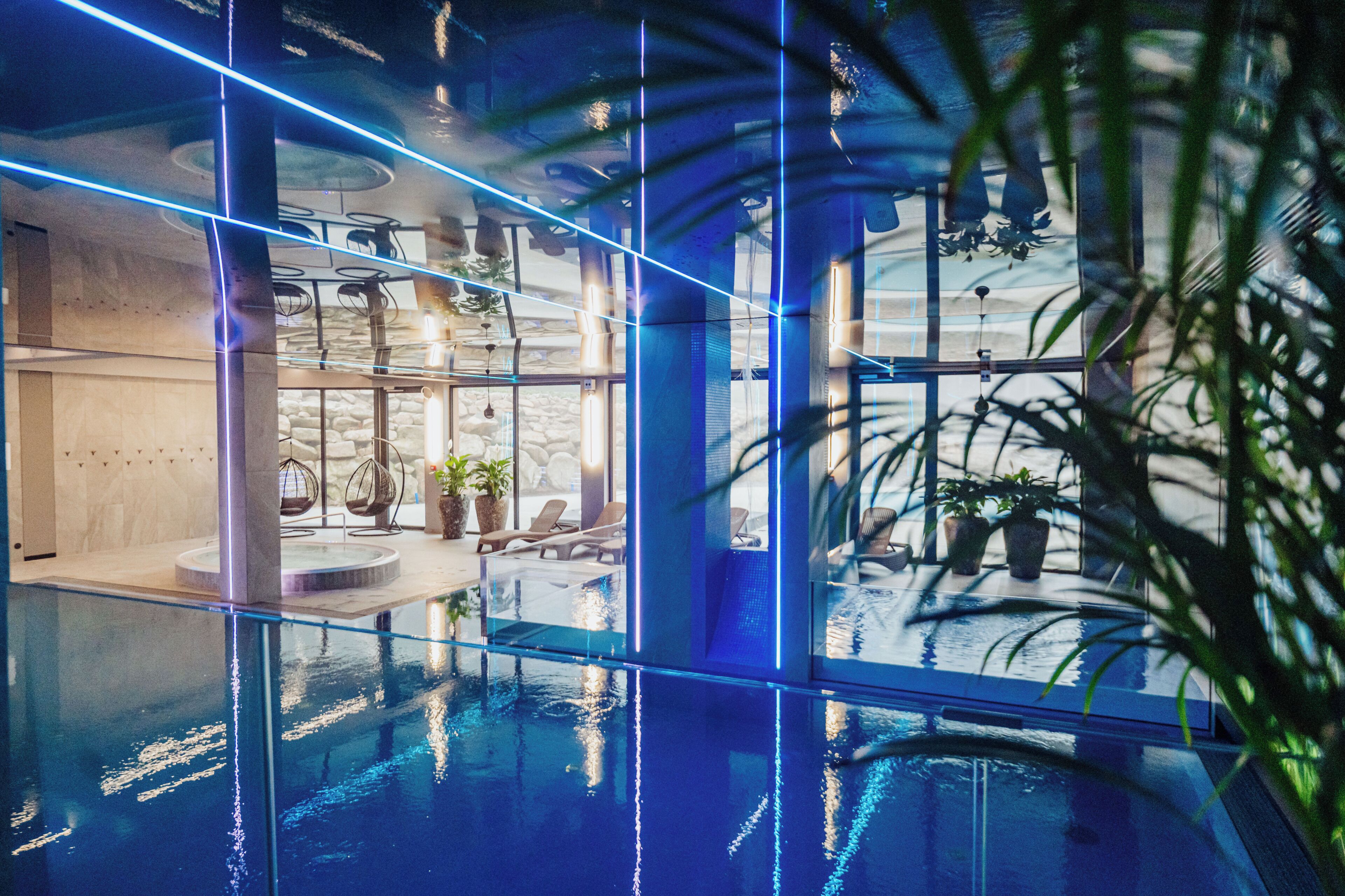 Uma piscina interior com paredes de vidro transparente, cadeiras de descanso e plantas em vasos.