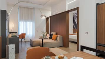 Quarto de hotel moderno com sofá, mesa de jantar com uma xícara e um doce, escrivaninha com cadeira e uma janela grande com cortinas.