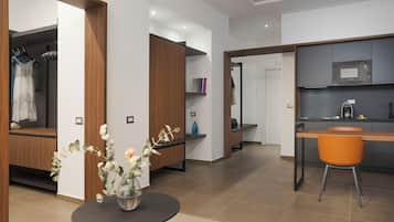 Quarto de hotel moderno com guarda-roupa de madeira, uma pequena mesa redonda com um vaso de flores, área de cozinha com micro-ondas e uma cadeira laranja.