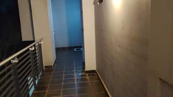 Um corredor com piso de azulejos, guarda-corpo de metal de um lado e luminária na parede.