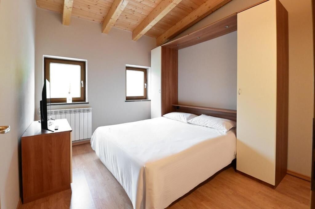 Quarto com cama, mesa de cabeceira de madeira, guarda-roupa e duas janelas.