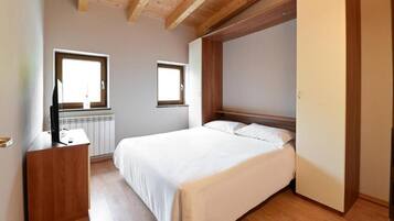Quarto com cama, mesa de cabeceira de madeira, guarda-roupa e duas janelas.