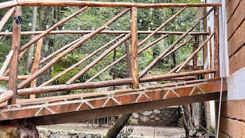 Uma ponte de madeira rústica com guarda-corpo de metal, que leva a um muro de pedra e um edifício com fachada de tijolos.