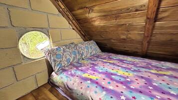 Um quarto pequeno e acolhedor com teto inclinado, paredes de madeira, uma cama simples com colcha estampada em estrelas e uma janela pequena com moldura decorativa.