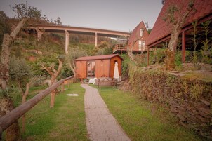 Uma cabana de madeira rústica com um caminho que a leva até ela, rodeada por vegetação e uma ponte ao fundo.