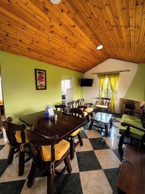 Uma sala de jantar com uma mesa e cadeiras de madeira, uma televisão na parede e uma janela com cortinas.
