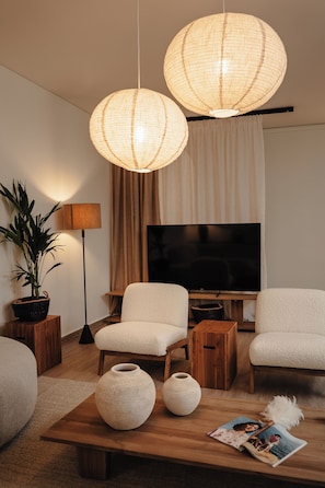 Uma sala de estar moderna com uma televisão de tela plana, duas poltronas brancas, uma mesa de centro com objetos decorativos e uma luminária trançada grande.