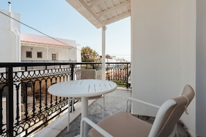 Uma varanda com mesa e cadeiras brancas, grade de ferro forjado preto e vista para edifícios ao fundo.