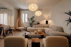 Uma sala de estar moderna com sofá, duas poltronas, uma mesa de centro e uma cadeira de ratã suspensa. Há um abajur em uma mesinha lateral e um vaso de planta.
