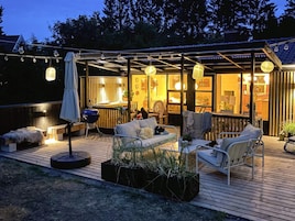 Área de estar externa com deck de madeira, assentos e luzes pendentes.