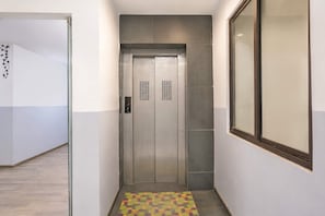 Um corredor estreito com chão ladrilhado, uma porta com visor digital e uma janela à direita.