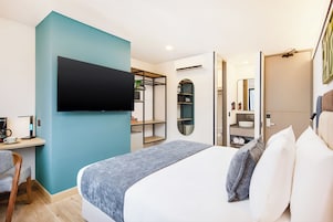 Quarto de hotel moderno com uma cama, uma escrivaninha com cafeteira, TV na parede e um guarda-roupa com prateleiras.