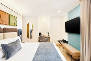 Quarto de hotel com cama, escrivaninha, cadeira, televisão e um banco.